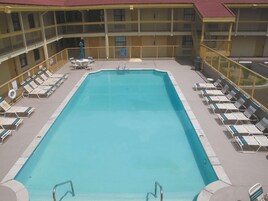Uma piscina retangular com um deck ladrilhado, rodeada por espreguiçadeiras e uma área de estar coberta.