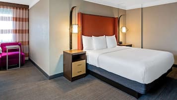 Quarto de hotel com uma cama grande, duas mesas de cabeceira, uma mesinha de apoio, uma cadeira e uma janela com cortinas.