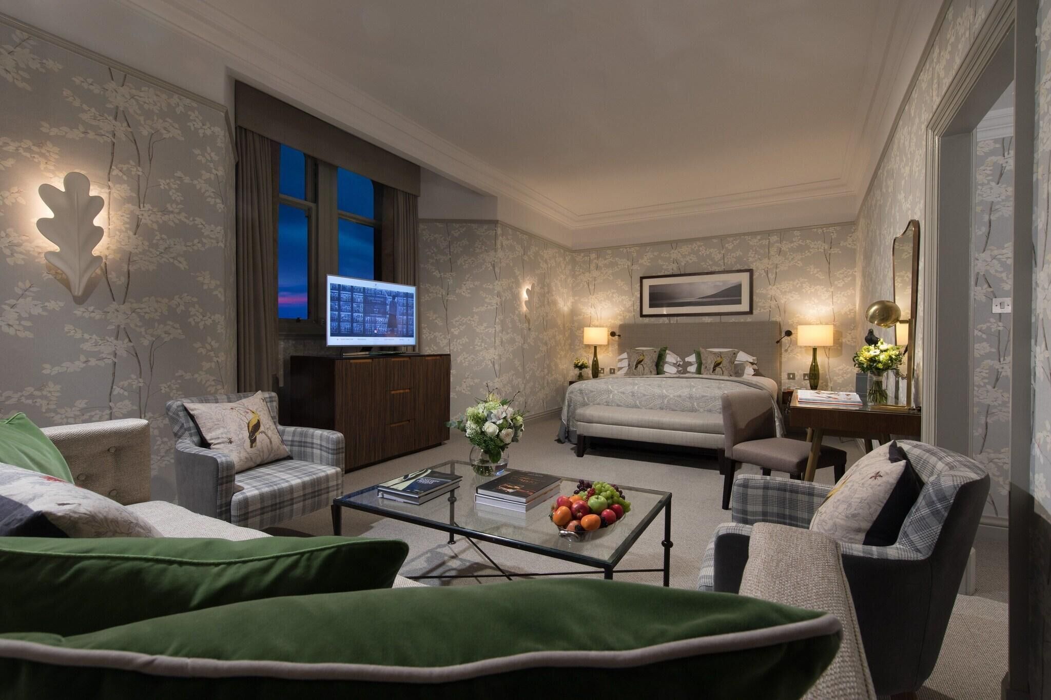 Quarto de hotel com uma cama, duas poltronas, um sofá, uma mesa de centro, uma televisão e uma escrivaninha com uma luminária e flores.