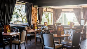 Interior de restaurante com mesas e cadeiras de madeira, grandes janelas com cortinas e uma parede de pedra.