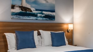 Quarto de hotel com cama, roupa de cama branca e azul, cabeceira de madeira e um quadro de um farol na parede.
