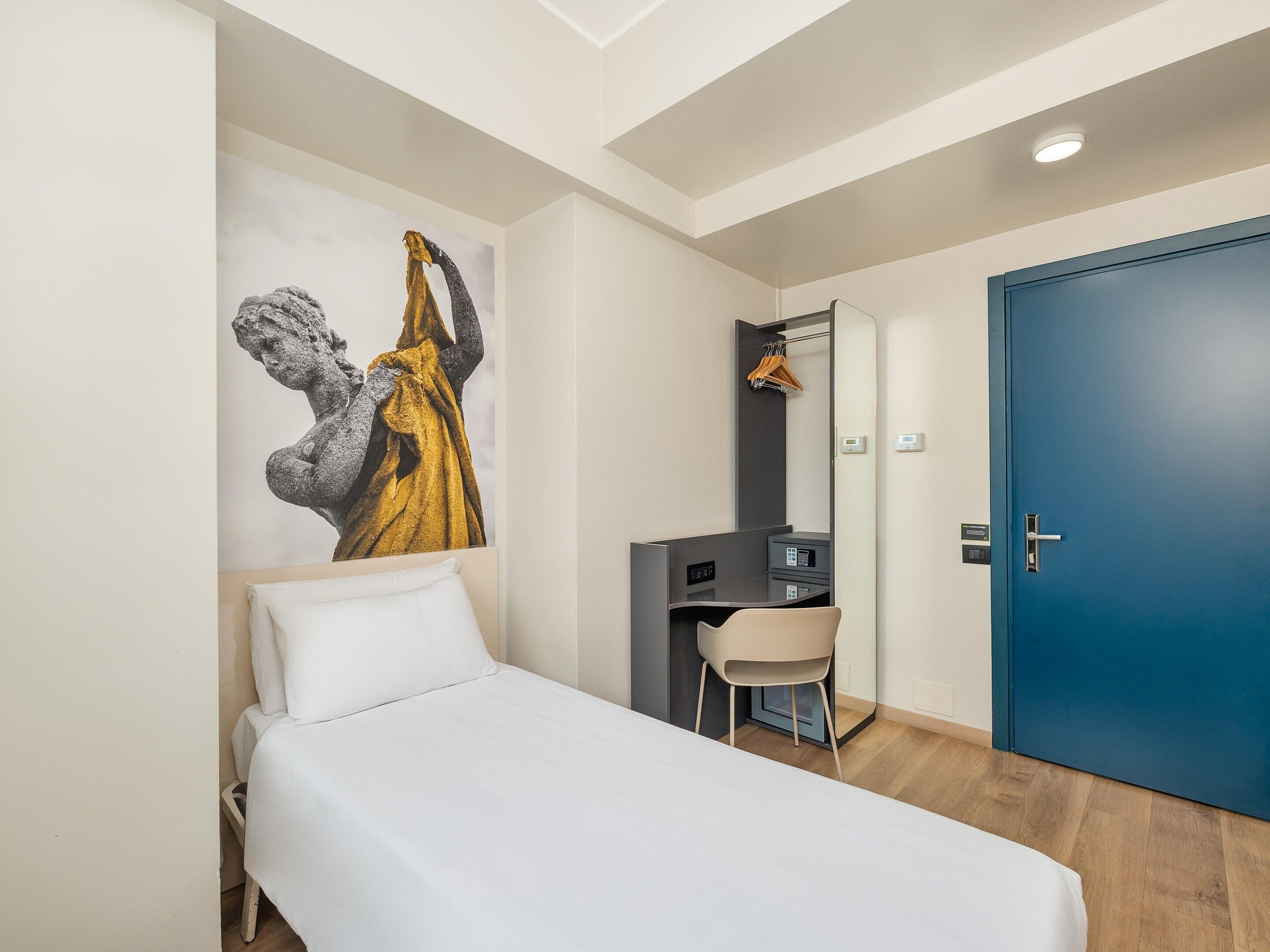 Quarto de hotel com cama, escrivaninha, cadeira, guarda-roupa e uma obra de arte pendurada na parede.