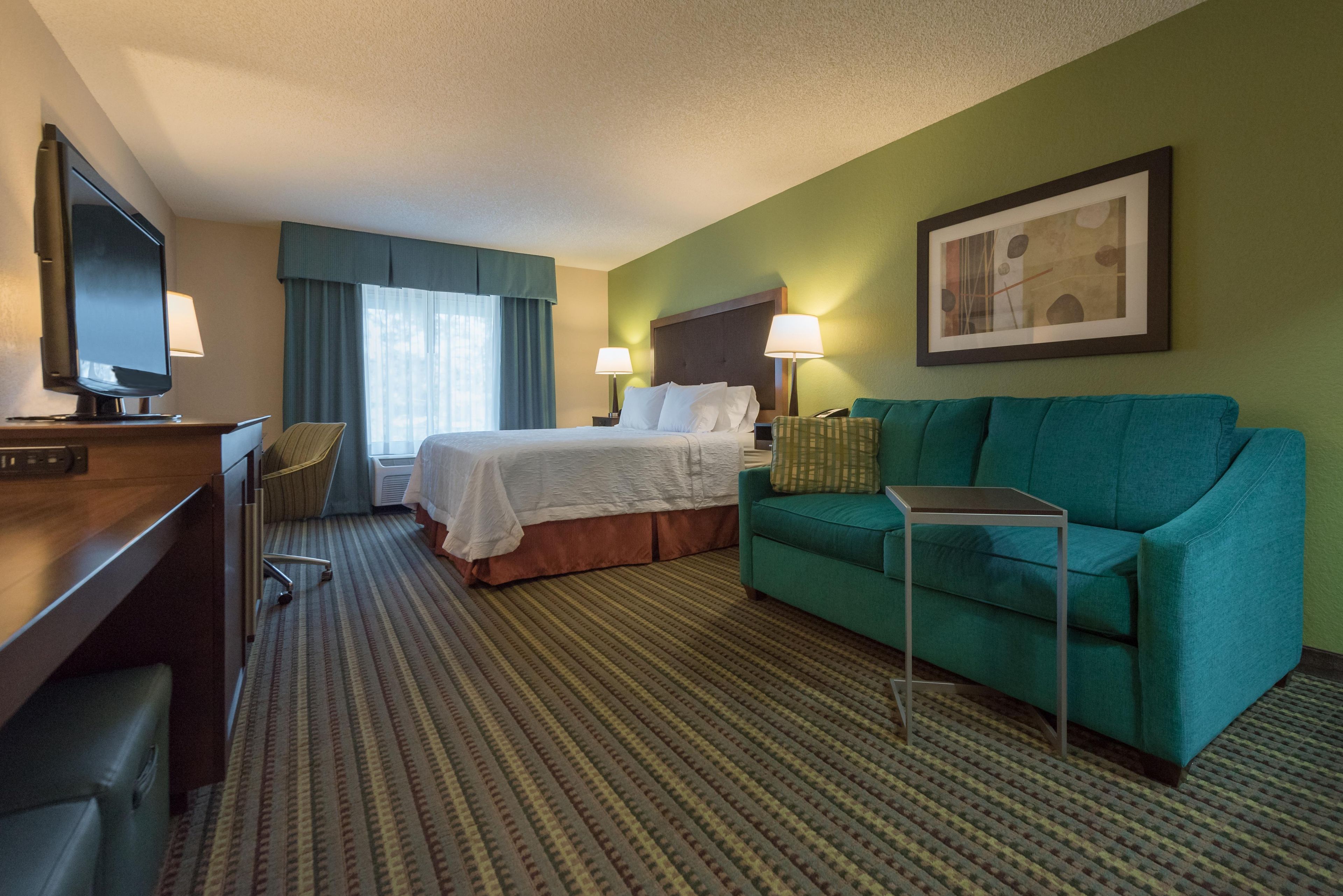 Quarto de hotel com cama, escrivaninha com televisão, sofá e quadro na parede.