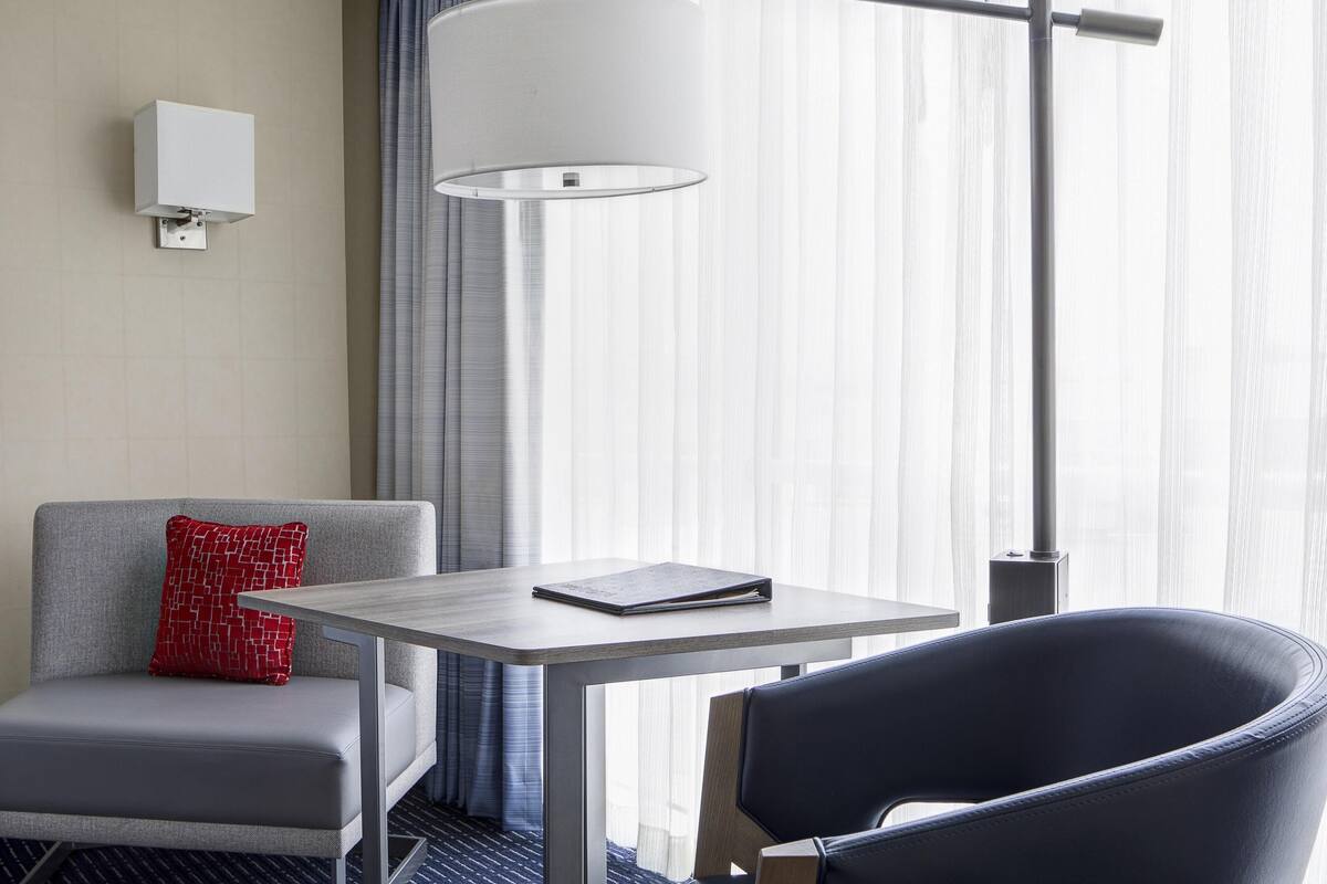 Quarto de hotel com sofá cinzento, almofada estampada vermelha, mesa pequena com um livro, luminária e vista através de cortinas leves.