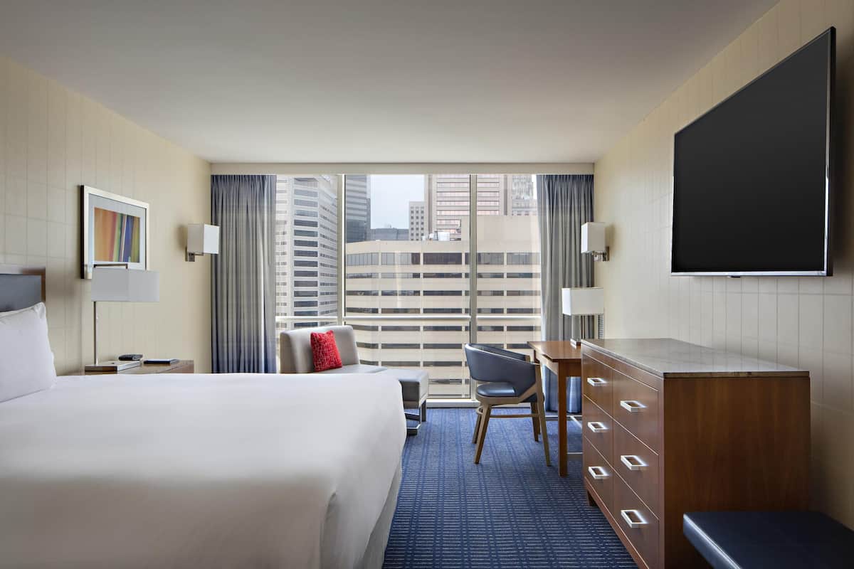 Quarto de hotel com cama, escrivaninha, cadeira, televisão e vista para edifícios.