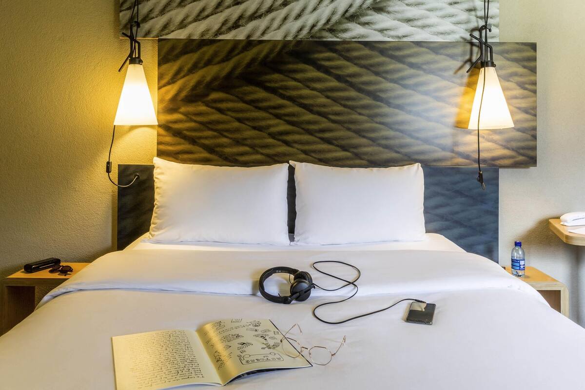 Cama bem arrumada com lençóis brancos, um par de fones de ouvido, um livro e um par de óculos sobre ela. Há duas mesas de cabeceira com luminárias, uma de cada lado da cama.