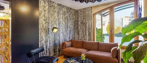 Uma sala de estar moderna com um sofá castanho, uma cadeira preta e uma mesa de centro redonda com um livro e um vaso. Apresenta duas luminárias de teto originais e uma janela ampla com vista para edifícios e montanhas.