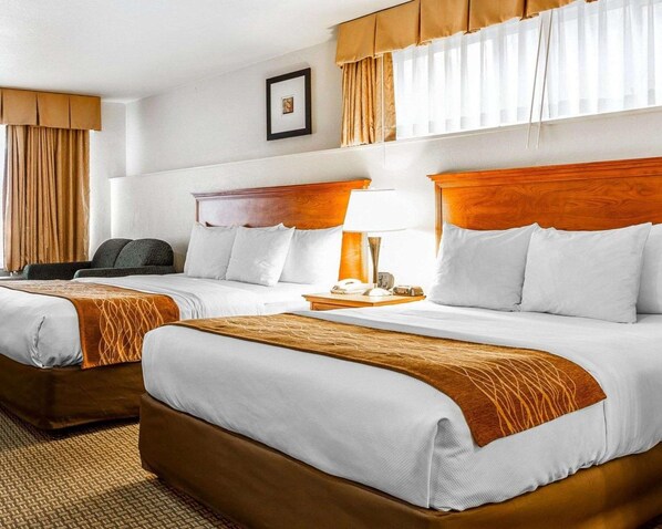 Quarto de hotel com duas camas, mesa de cabeceira com abajur, quadro na parede e janela com cortinas.