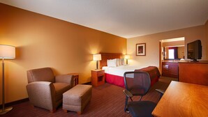 Quarto de hotel com uma cama, dois poltronas, uma escrivaninha, uma cadeira, uma TV e um banheiro visível através de uma porta aberta.