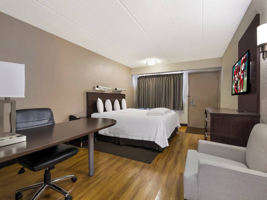 Quarto de hotel com cama, escrivaninha, cadeira e televisão na parede.