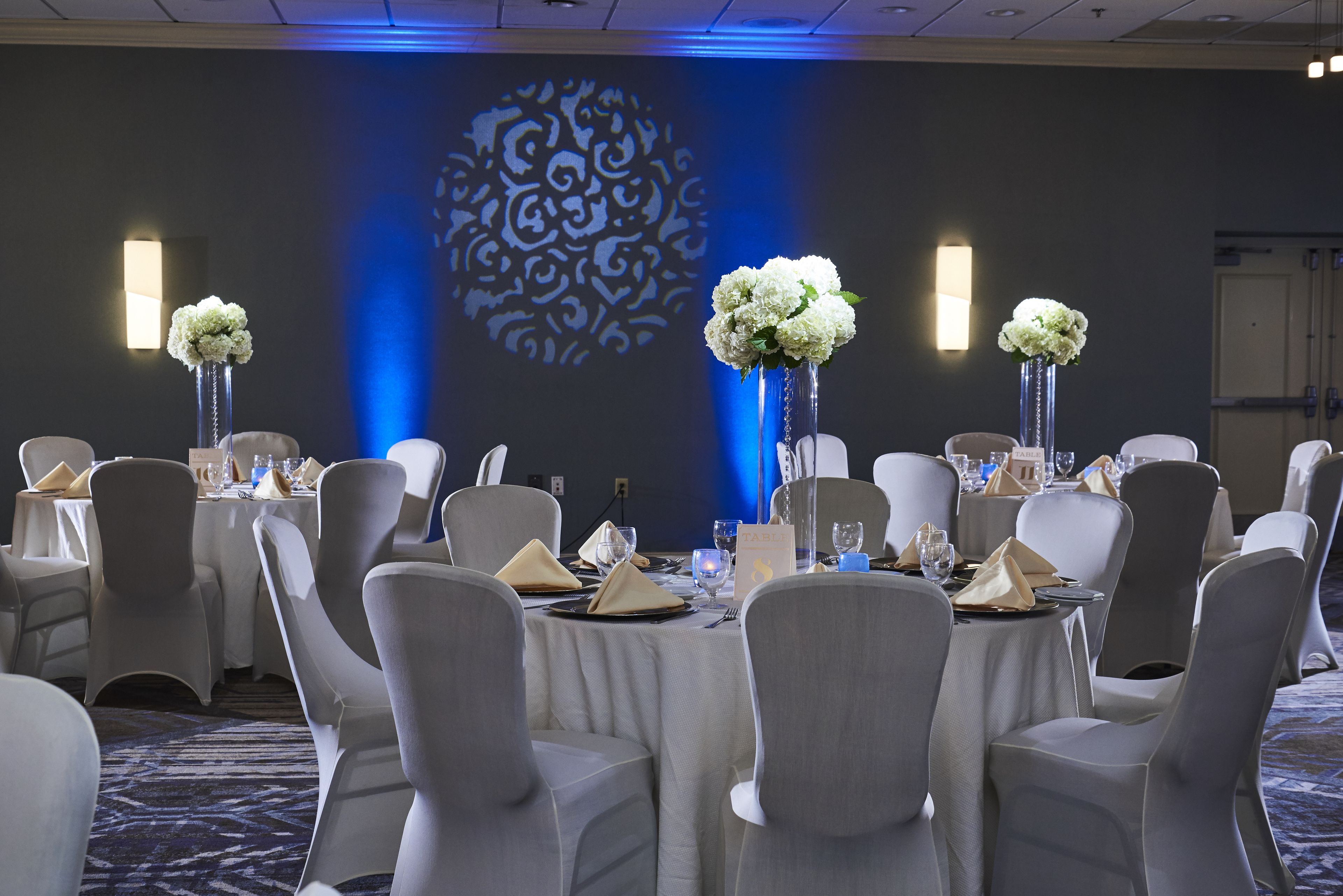 Uma sala de banquetes com mesas redondas, cadeiras brancas e centros de mesa florais.