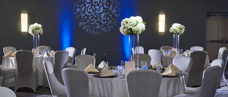 Uma sala de banquetes com mesas redondas, cadeiras brancas e centros de mesa florais.