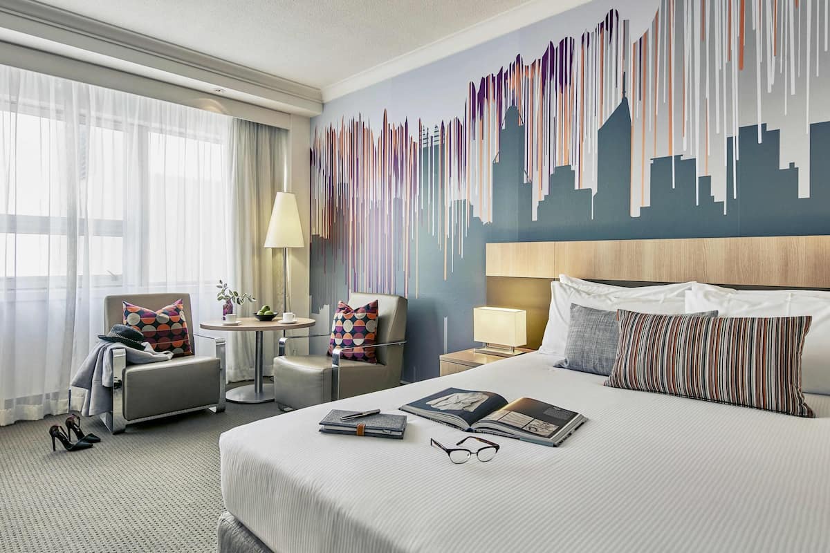 Quarto de hotel com uma cama grande, duas poltronas, uma mesa pequena e um mural com vista para a cidade na parede.
