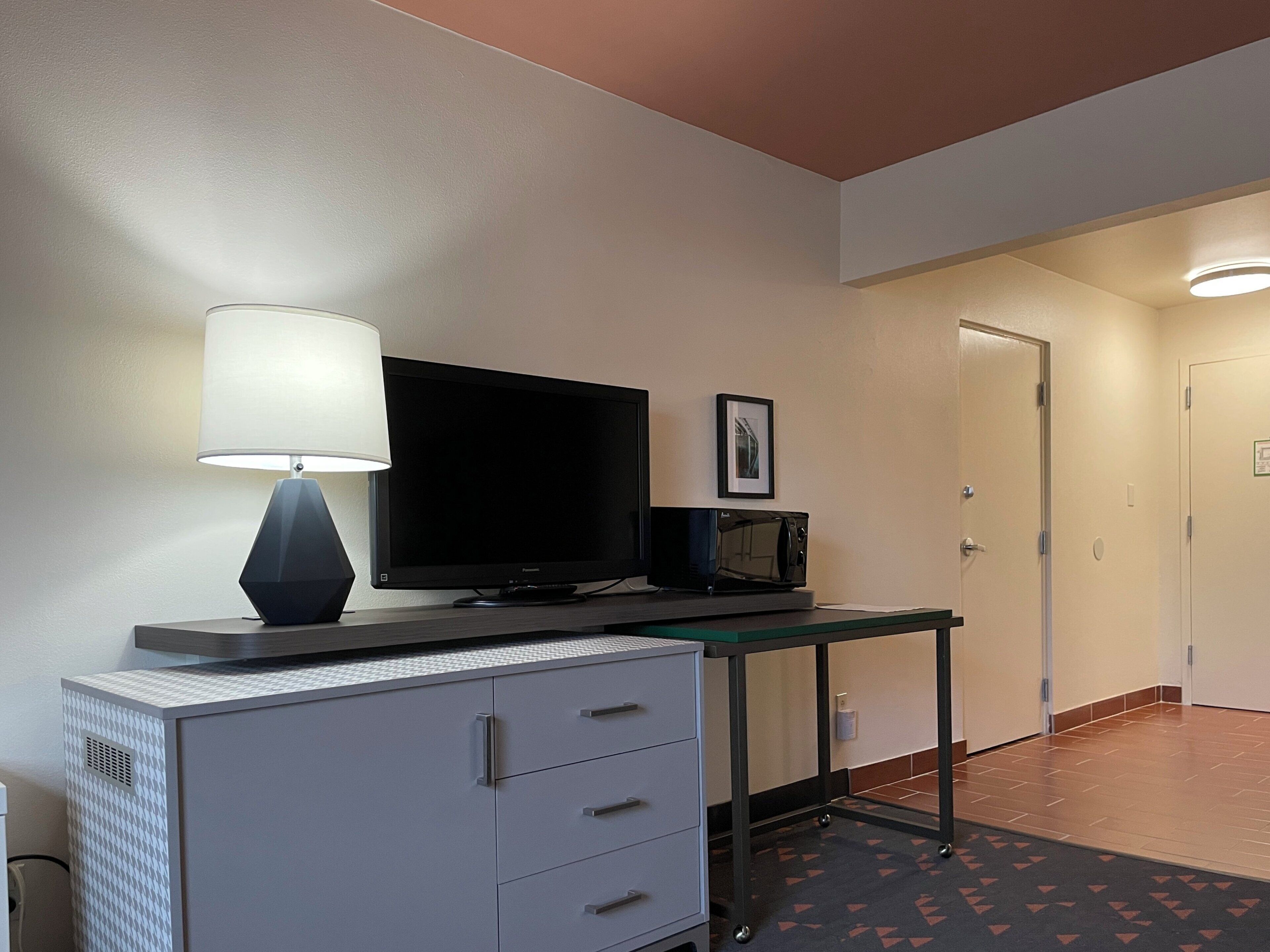 Quarto de hotel com escrivaninha, televisão, micro-ondas, armário e porta.