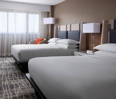 Quarto de hotel com duas camas, uma cadeira, uma mesinha de cabeceira e uma luminária.