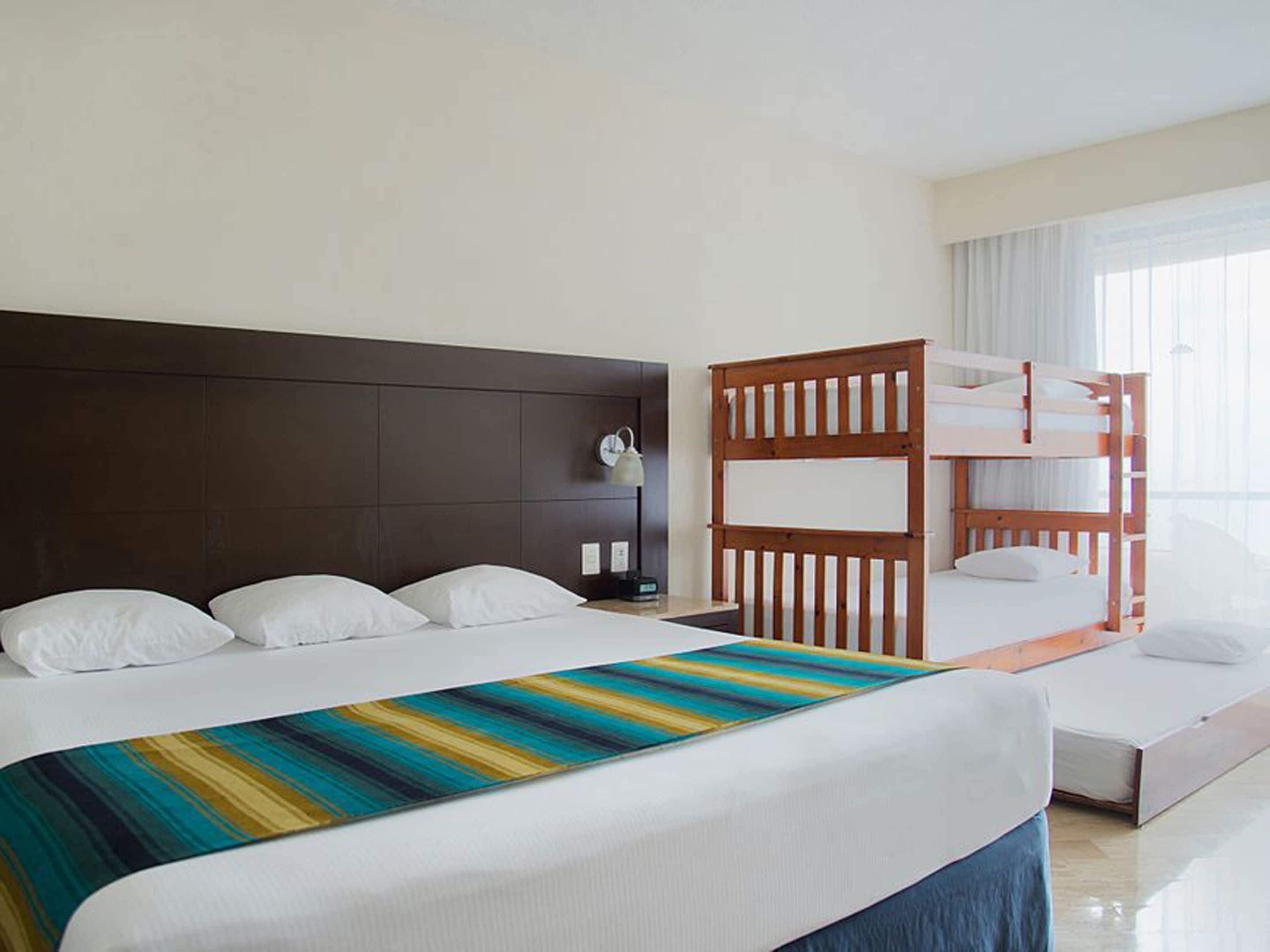Quarto de hotel com cama grande, beliche, mesa de cabeceira com abajur e janela com cortinas.