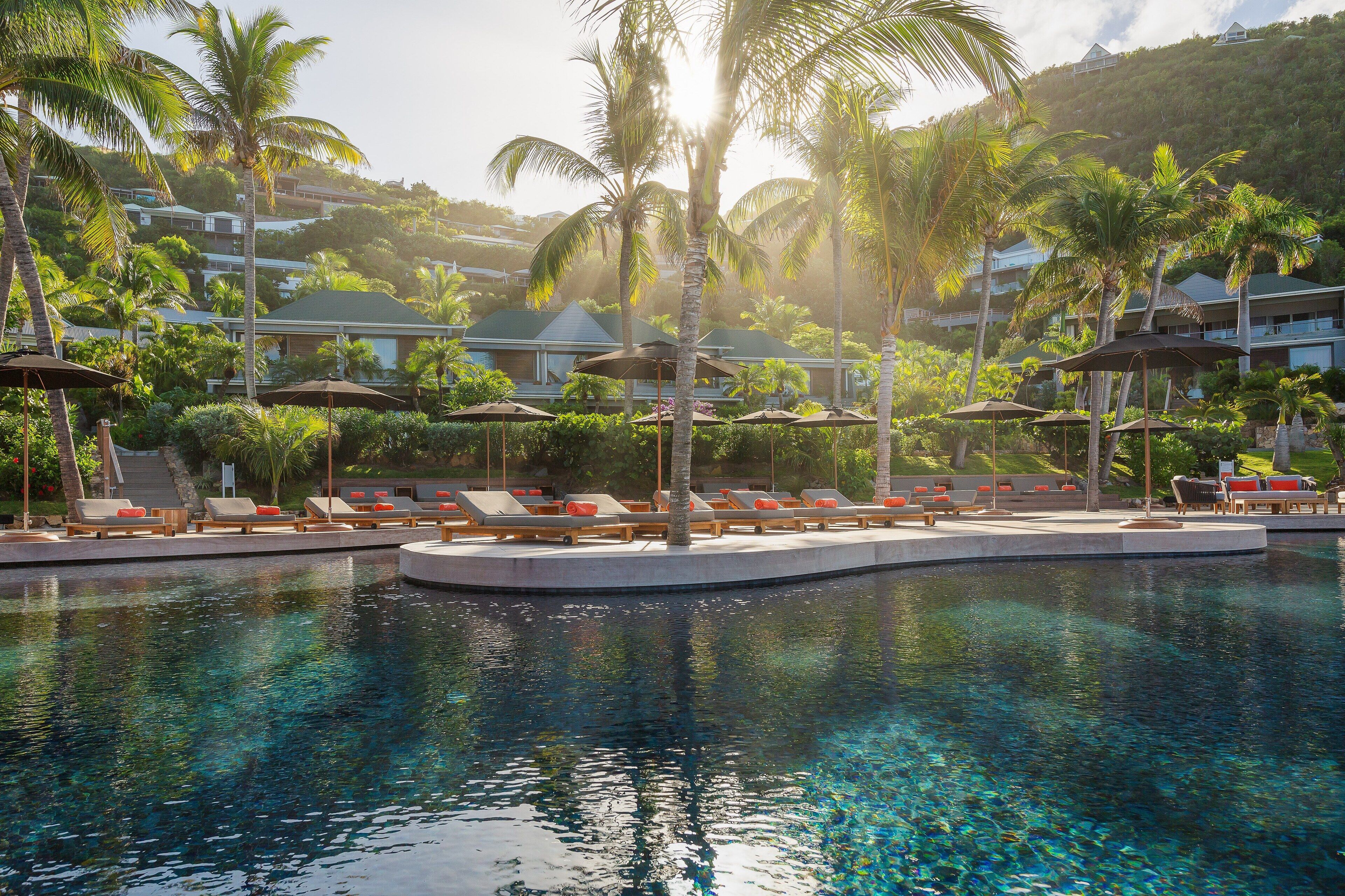 Piscina de resort com espreguiçadeiras e guarda-sóis, cercada por palmeiras e edifícios.