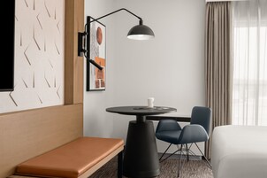 Quarto de hotel com cama, um banco, uma mesa redonda com cadeiras, uma luminária na parede e um quadro emoldurado.