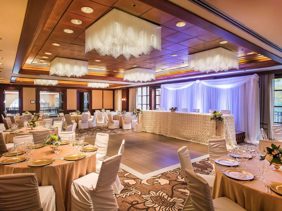 Salão de banquetes com mesas redondas preparadas para um evento formal, com um candelabro e cortinas com drapeado.