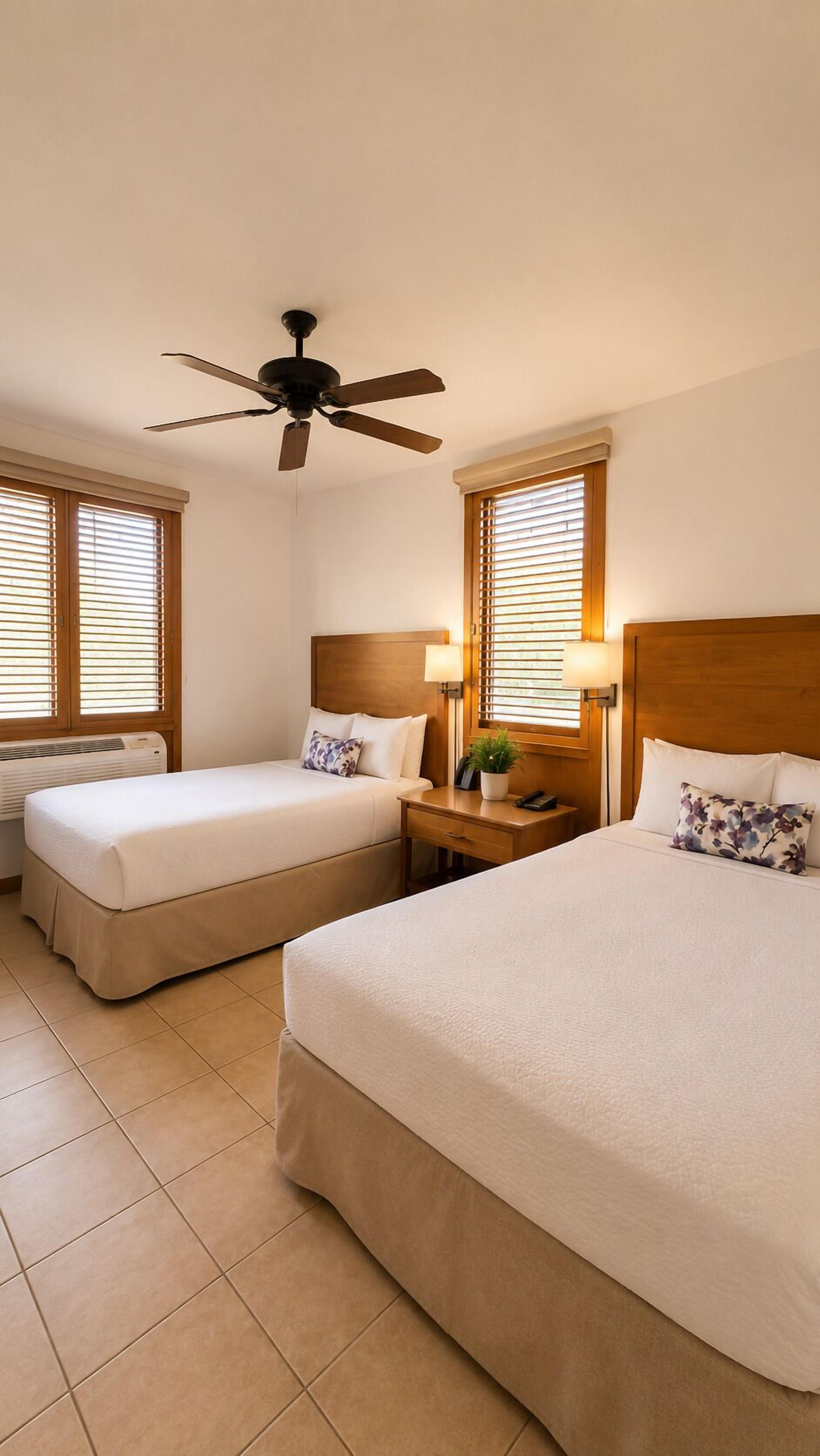 Quarto de hotel com duas camas, um ventilador de teto, uma mesa de cabeceira com abajur e uma janela com persianas.