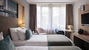 Quarto de hotel com duas camas, uma escrivaninha, uma cadeira, uma televisão e uma janela grande com cortinas.