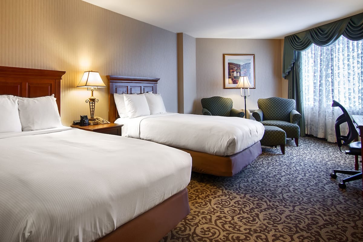 Quarto de hotel com duas camas, uma escrivaninha, uma cadeira e um abajur.