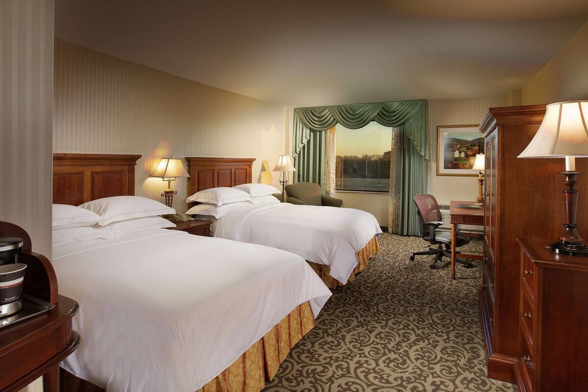 Quarto de hotel com duas camas, uma escrivaninha, uma cadeira e um abajur.