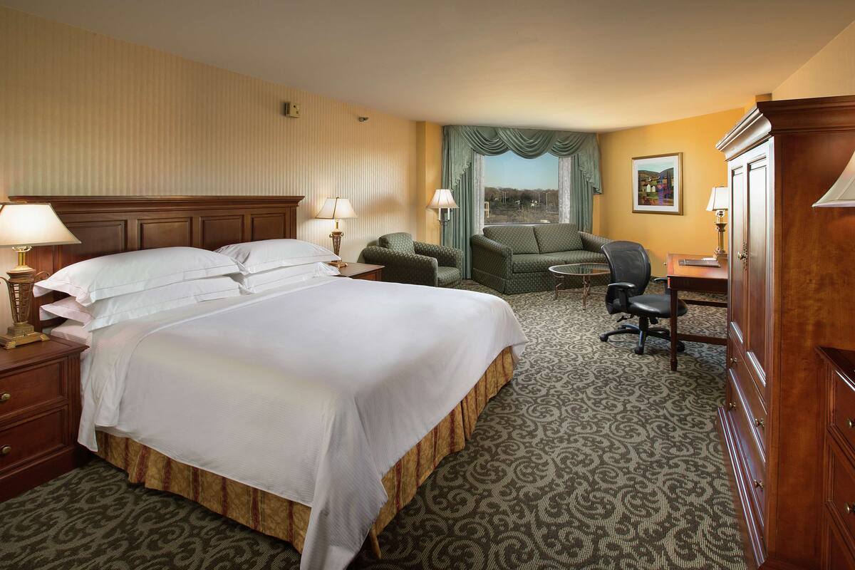 Quarto de hotel com uma cama grande, duas cadeiras, uma escrivaninha e vista para o exterior.