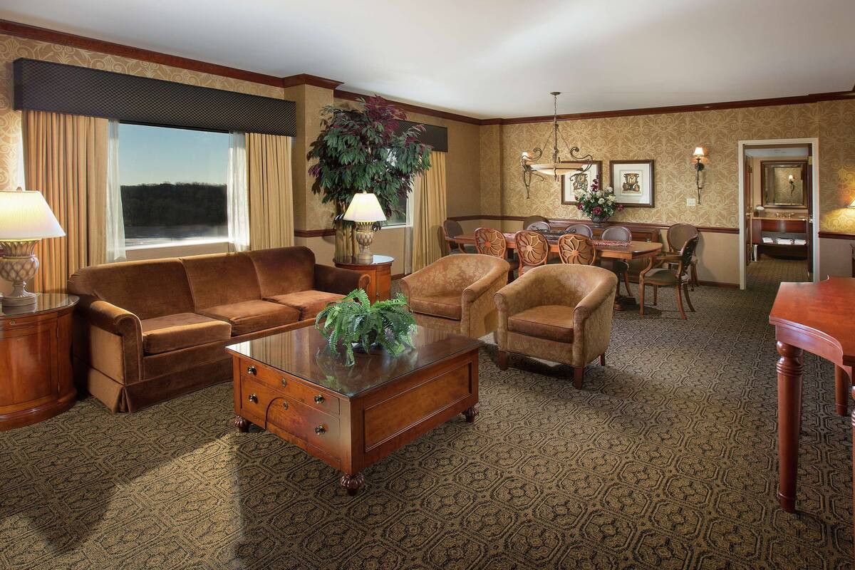 Quarto de hotel com área de estar, incluindo sofá, poltronas, mesa de centro e mesa de jantar com cadeiras. Há uma janela grande com cortinas, uma planta em vaso e um candelabro.