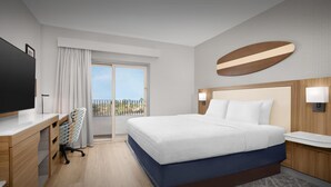 Quarto de hotel com cama, escrivaninha, televisão, cadeira, varanda e luminária na parede.