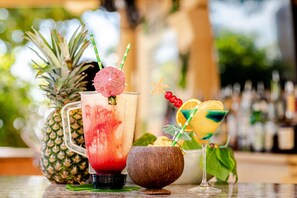 Um bar com abacaxi, um copo alto de bebida com canudo, coco e um coquetel com guarnição de frutas.