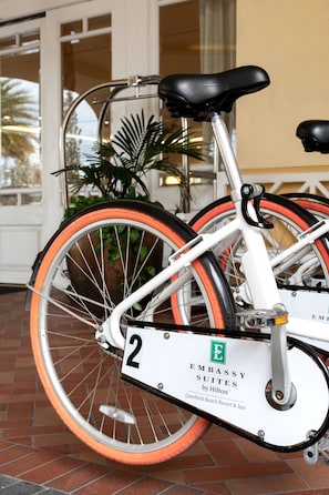 Uma bicicleta com estrutura branca e rodas laranja, estacionada em frente a um edifício com porta de vidro e uma placa que diz "Embassy Suites by Hilton Deerfield Beach Resort & Spa".