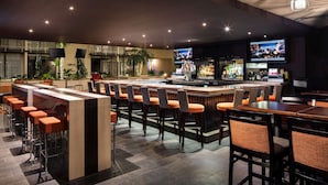Uma área de bar com banquetas altas, um balcão e uma tela de televisão.