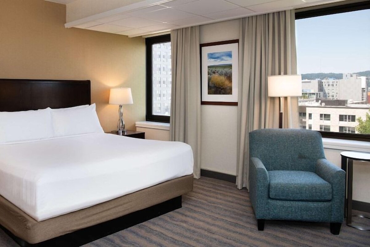 Quarto de hotel com uma cama grande, duas luminárias de cabeceira, uma cadeira, uma janela com vista para edifícios e um quadro na parede.