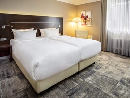 Uma cama de casal com roupa de cama branca, cabeceira de madeira, mesa de cabeceira, luminária e um quadro na parede.