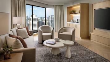 Quarto de hotel moderno com sofá, duas poltronas, mesa de centro, televisão de tela plana grande, área de cozinha e vista para a paisagem urbana através de amplas janelas.