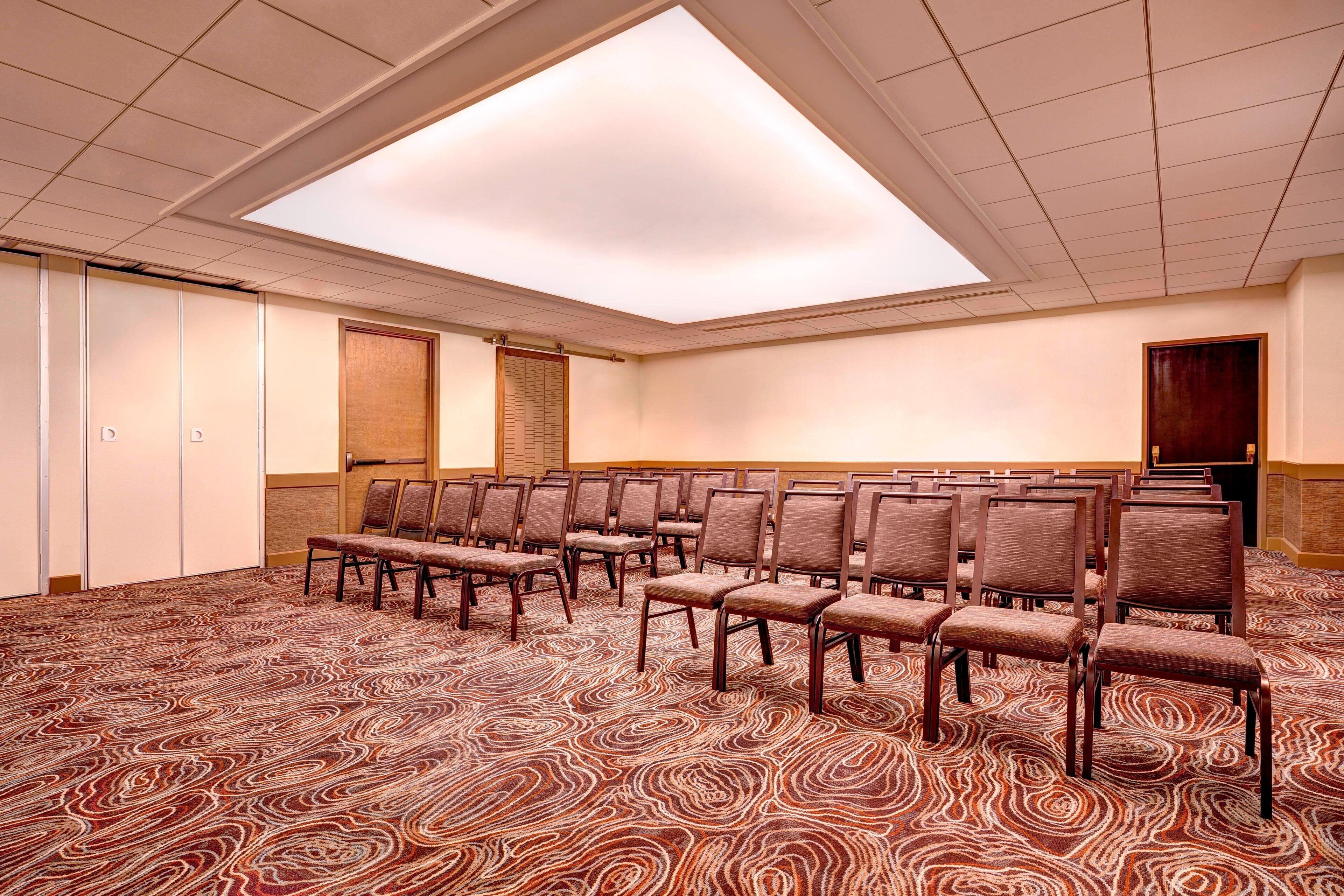 Uma sala de conferências com fileiras de cadeiras, um tapete estampado, um claraboia e uma porta de madeira.