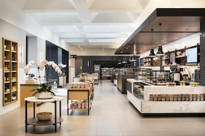 Interior moderno de café com balcão central, prateleiras com bebidas e área de estar com mesas e cadeiras.