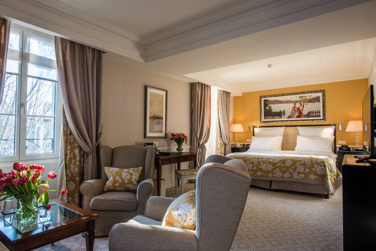 Quarto de hotel com cama, duas poltronas, uma mesinha de centro com um vaso de flores, escrivaninha e um quadro na parede.