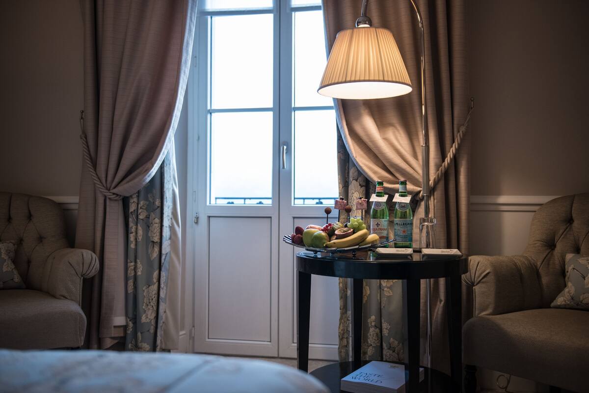 Quarto de hotel com uma cama, duas poltronas, uma mesa com frutas e bebidas, e uma janela com cortinas.