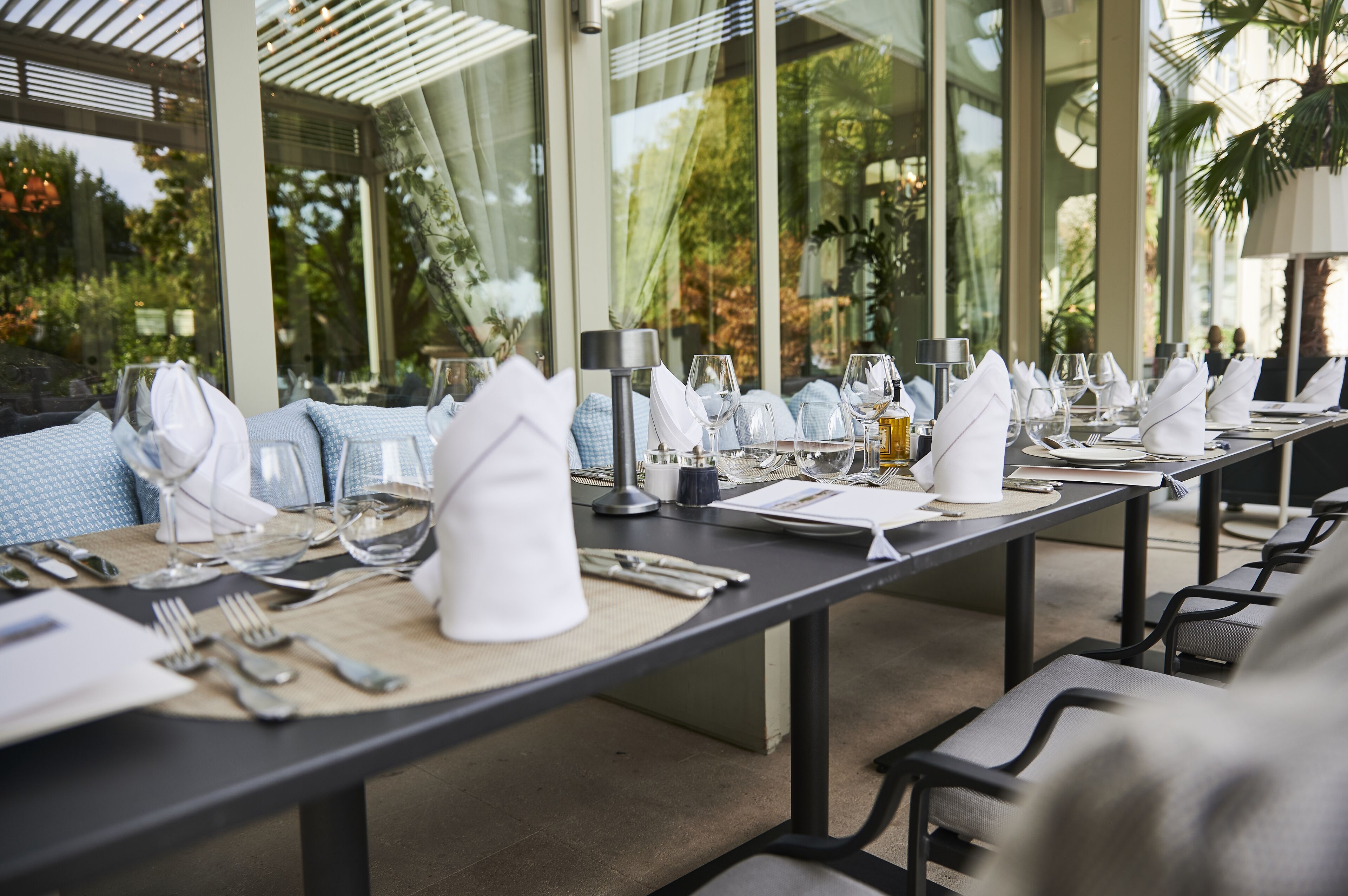 Mesa de jantar posta com guardanapos brancos, talheres e copos, com vista para o verde através de grandes janelas.