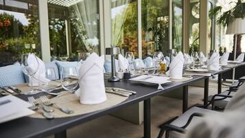 Mesa de jantar posta com guardanapos brancos, talheres e copos, com vista para o verde através de grandes janelas.