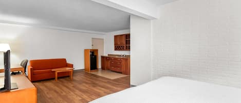Quarto de hotel com cama, escrivaninha com computador, sofá laranja e cozinha com armários de madeira.