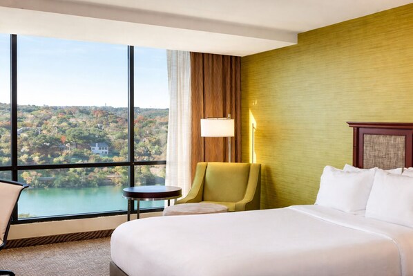 Quarto de hotel com uma cama grande, uma poltrona verde, uma mesa pequena e vista para um lago e colinas.