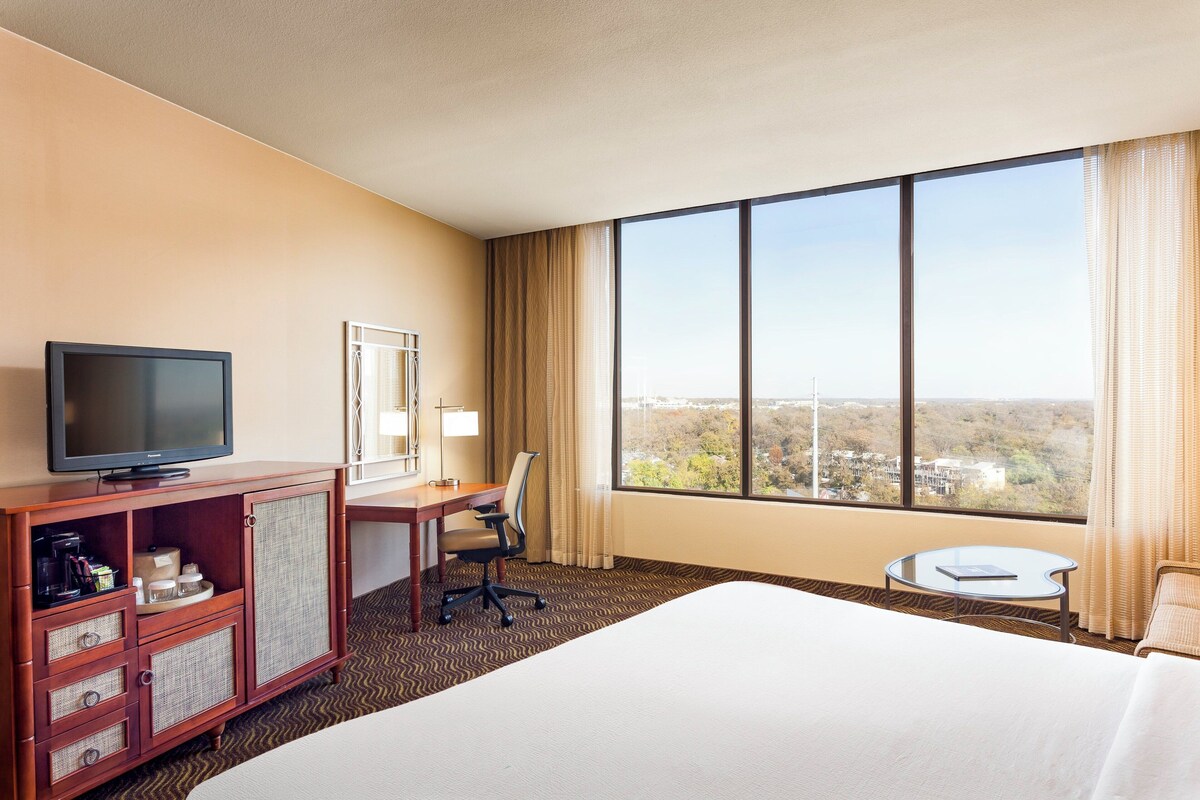 Quarto de hotel com uma cama grande, uma mesa com cadeira, televisão, uma mesinha e janela com vista para edifícios.