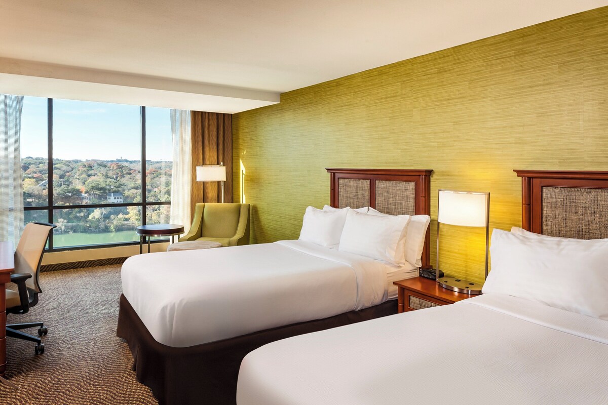 Quarto de hotel com duas camas, uma escrivaninha, uma cadeira e uma janela ampla com vista para o exterior.