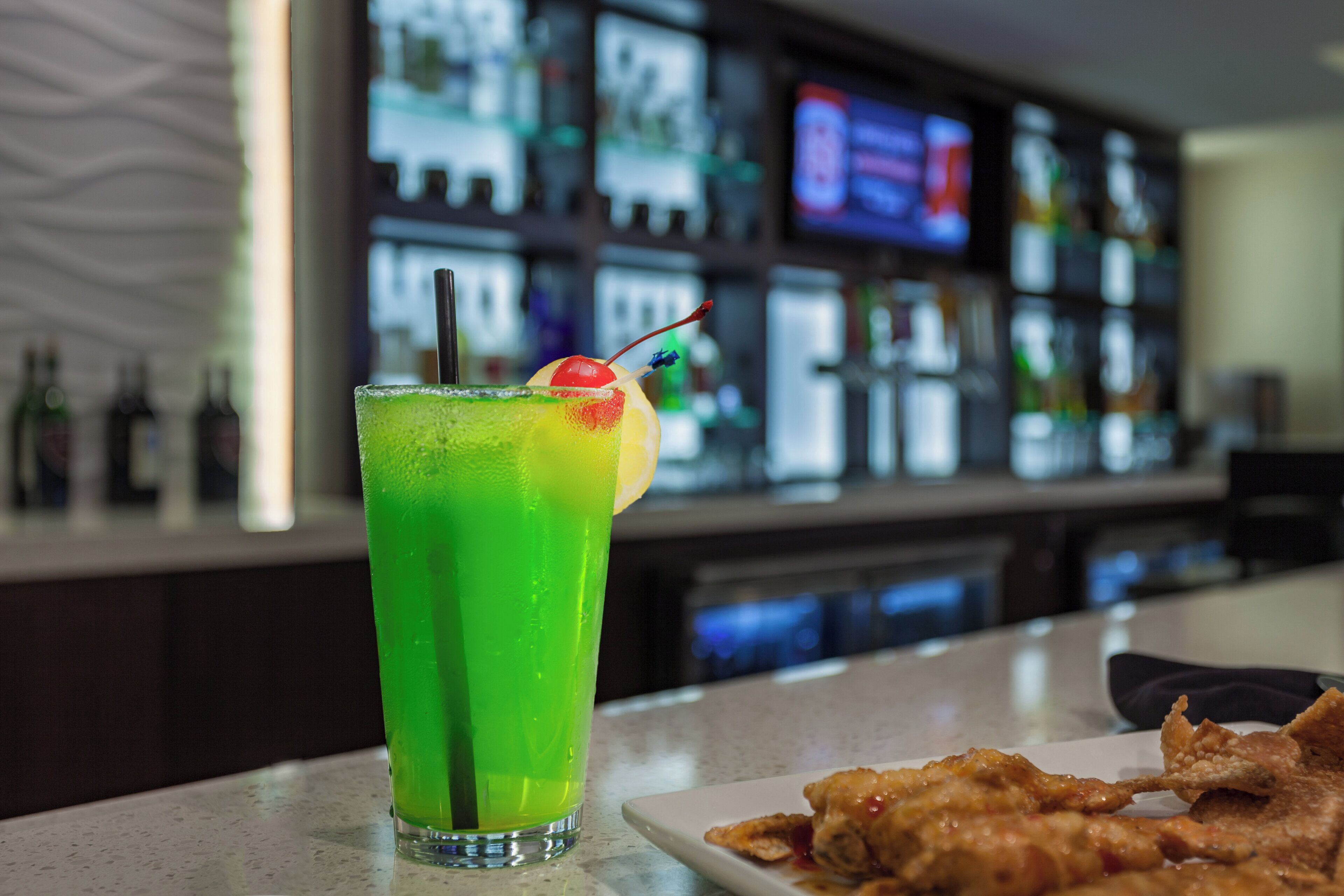 Ambiente de bar com um cocktail verde, um prato de comida frita e um balcão com prateleiras de garrafas.