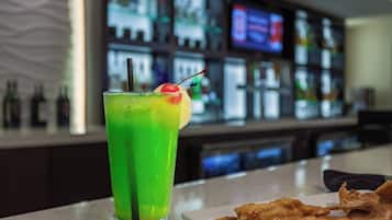 Ambiente de bar com um cocktail verde, um prato de comida frita e um balcão com prateleiras de garrafas.