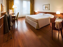 Quarto de hotel com uma cama grande, duas luminárias de cabeceira, uma escrivaninha com luminária, uma cadeira e um cabideiro.