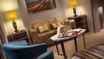 Quarto de hotel moderno com sofá, poltrona azul, mesa de jantar com bandeja de petiscos e um quadro na parede.